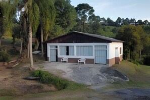 Um edifício térreo com uma grande porta de garagem, um alpendre com duas cadeiras e um caminho de acesso de terra.