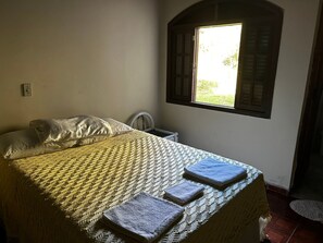 Uma cama de solteiro com colcha e almofadas estampadas, uma mesinha de cabeceira com abajur e uma janela com persianas.
