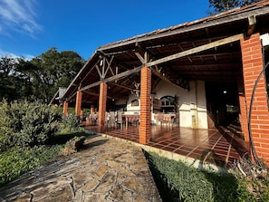 Área externa coberta com colunas de tijolo, piso de pedra e um pátio com mesas e cadeiras.