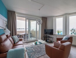 Uma sala de estar moderna com um sofá, poltronas, uma mesa de centro e uma televisão. Há uma porta de vidro deslizante que dá para uma varanda com vista para o mar.