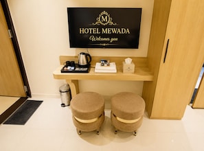 Quarto de hotel com televisão exibindo o nome do hotel, uma pequena mesa com bandeja de amenidades, dois pufes e uma lixeira.