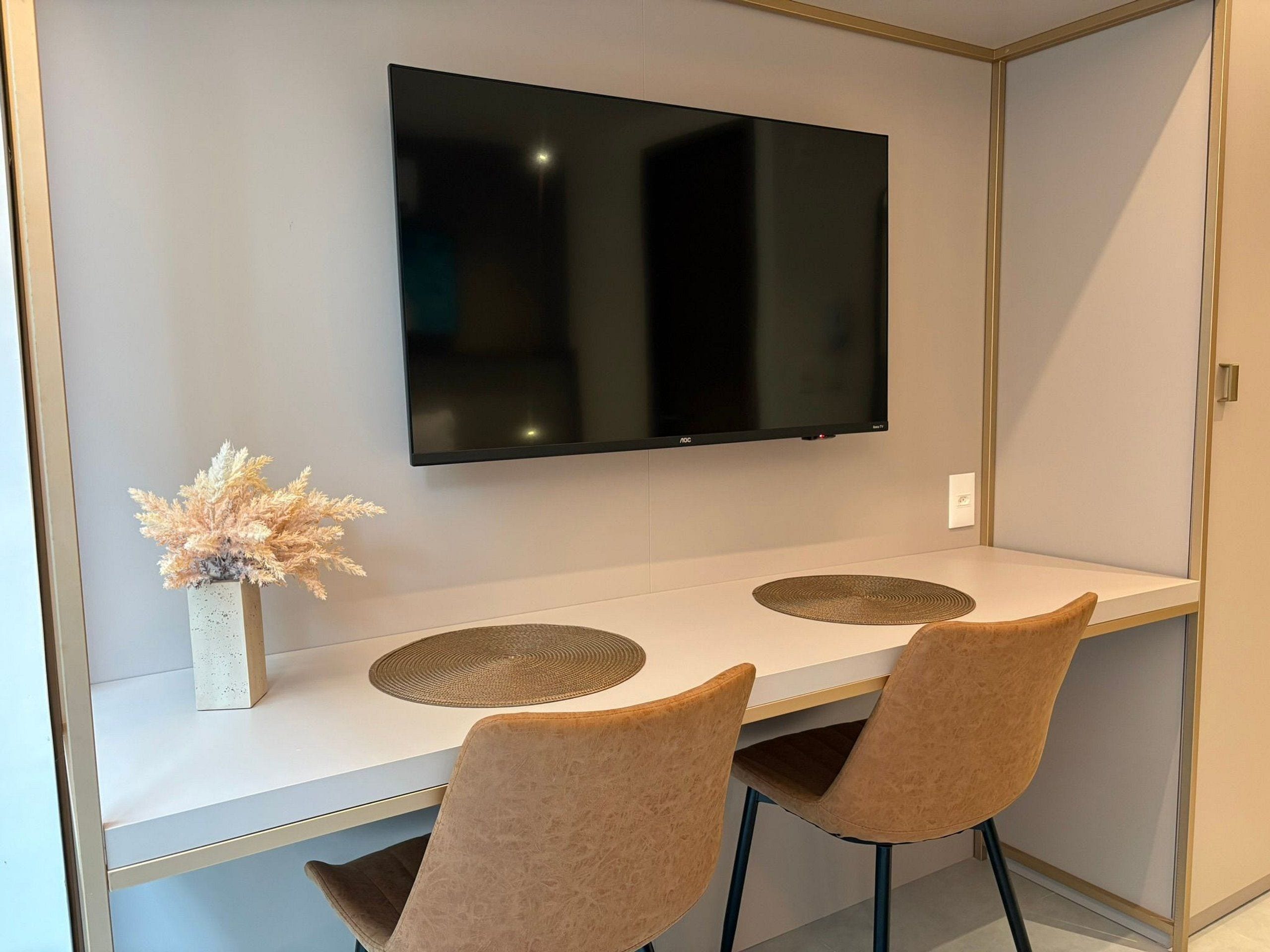 Uma área de jantar moderna com uma televisão de tela plana na parede, uma mesa branca, duas cadeiras e um vaso decorativo com flores secas.