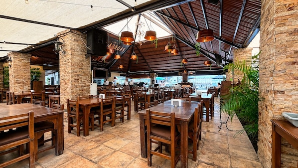 Um restaurante com mesas e cadeiras de madeira, pilares de pedra, luminárias pendentes e vista para a rua.
