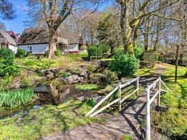 Uma casa rústica com um lago, uma ponte de madeira e vegetação ao redor.