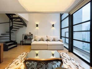 Uma sala de estar moderna com um sofá, uma mesa de centro de vidro, um tapete estampado, uma escada e uma grande janela com vista para a cidade.