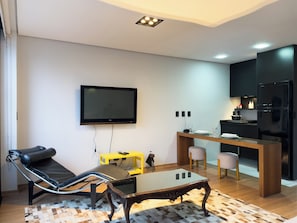 Uma sala de estar moderna com televisão de tela plana, sofá, mesa de centro e uma área de cozinha com geladeira e fogão.