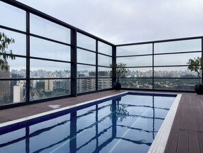 Piscina no terraço com vista para a cidade, cercada por paredes de vidro.
