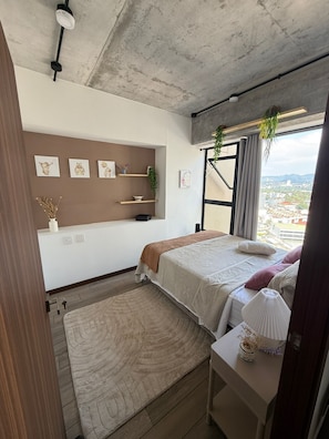 Quarto com duas camas, janela com vista para a cidade, prateleira na parede com objetos decorativos e luminária no teto.