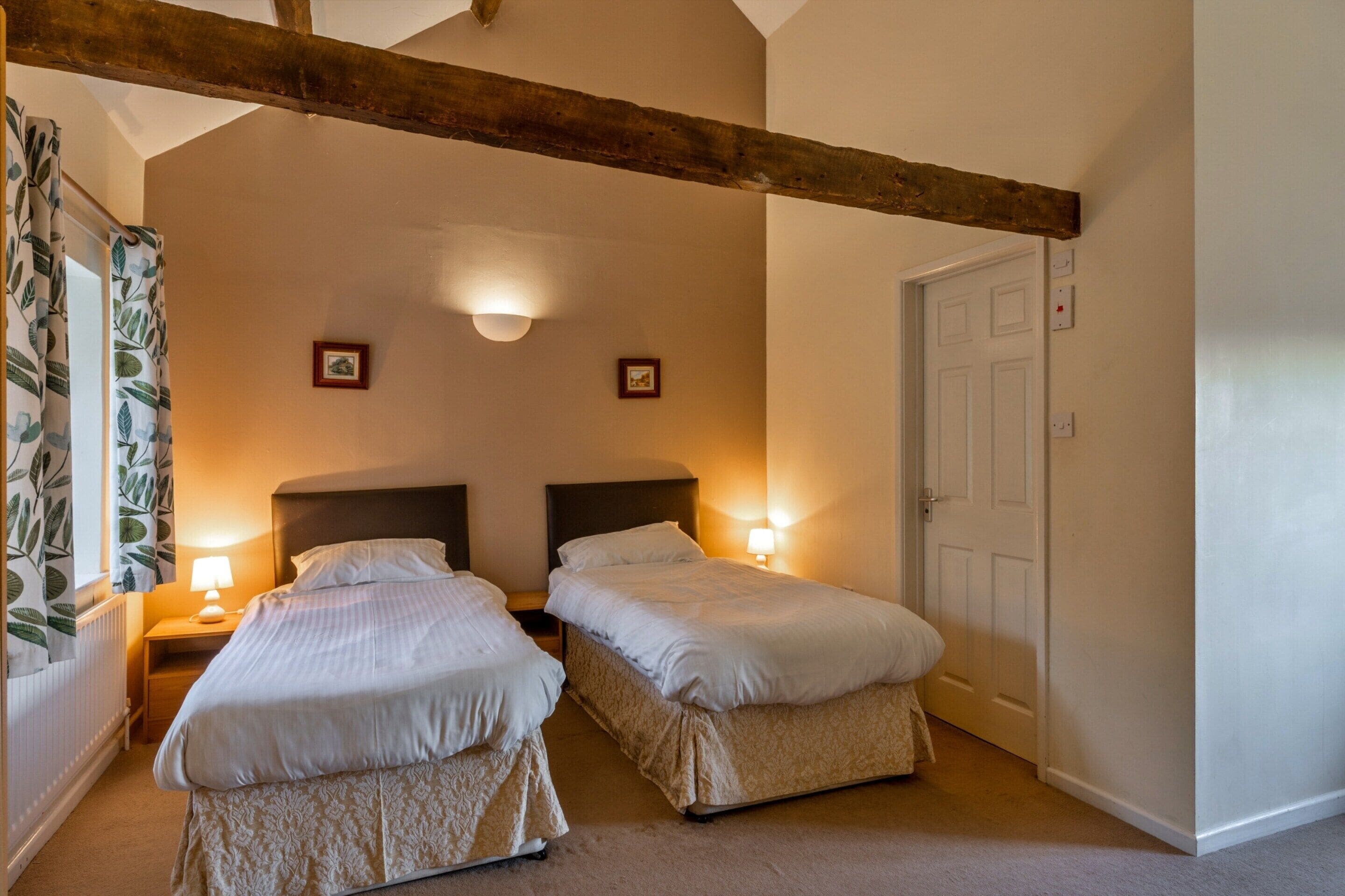 Quarto de hotel com duas camas, uma viga de madeira no teto, uma porta e duas luminárias fixadas na parede.