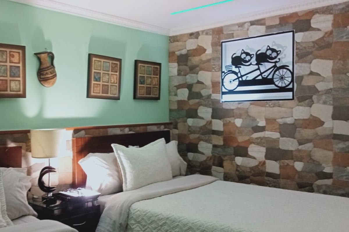 Quarto com cama, mesa de cabeceira, luminária e parede com padrão de pedra e quadros emoldurados.