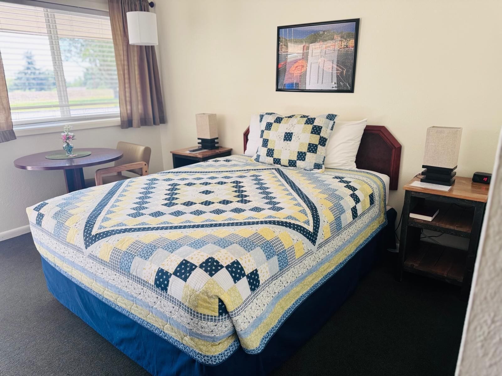Quarto de hotel com uma cama grande, uma mesa de cabeceira com abajur, uma mesinha com um vaso de flores e um quadro na parede.