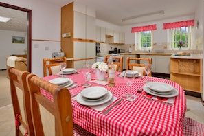 Uma sala de jantar com toalha xadrez vermelha e branca, cadeiras de madeira e uma cozinha ao fundo.