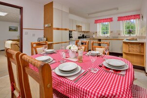 Uma sala de jantar com toalha xadrez vermelha e branca, cadeiras de madeira e uma cozinha ao fundo.