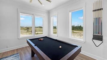 Uma mesa de bilhar em um espaço amplo com janelas grandes e um ventilador de teto.