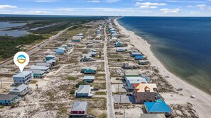 Uma área costeira com uma fileira de casas de praia ao longo da linha da costa, uma estrada correndo paralelamente à praia, e um corpo de água ao longe.