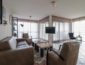 Uma sala de estar com sofá, cadeiras, mesa de centro, televisão e uma janela grande com cortinas.