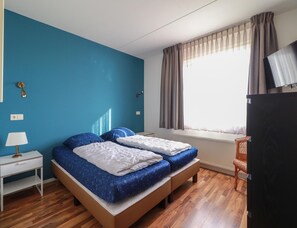 Quarto com cama, mesa de cabeceira, abajur, janela com cortinas e televisão.