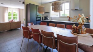 A modern dining area with a large table and chairs, a kitchen in the background, and a window with a view.