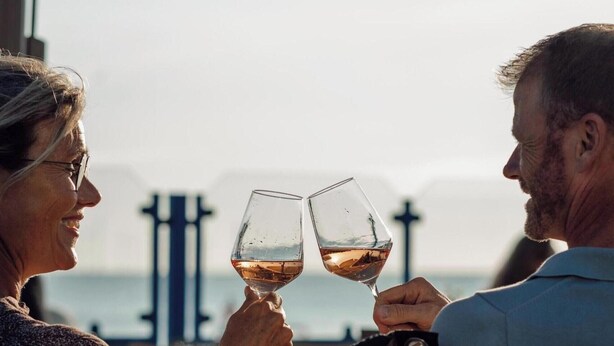Two people toasting with wine glasses at a seaside table with a view of the sea and a pier in the background.