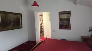 Quarto com cama, abajur vermelho, um quadro e uma porta que dá para outro cômodo.