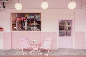 Paredes externas cor de rosa com uma janela mostrando o letreiro de uma cafeteria, duas cadeiras cor de rosa e uma porta.