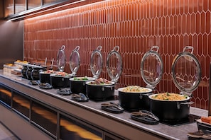 Buffet com diversos pratos em panelas pretas, algumas tampadas, em frente a uma parede de azulejos vermelhos.