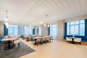 Uma sala de jantar moderna com várias mesas e cadeiras, janelas amplas e teto alto com luminárias pendentes.