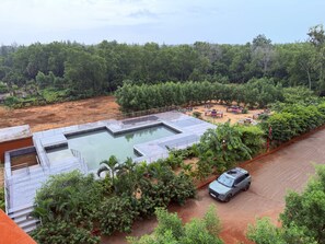 Piscina com um deck ao redor, um veículo estacionado nas proximidades e uma área arborizada ao fundo.