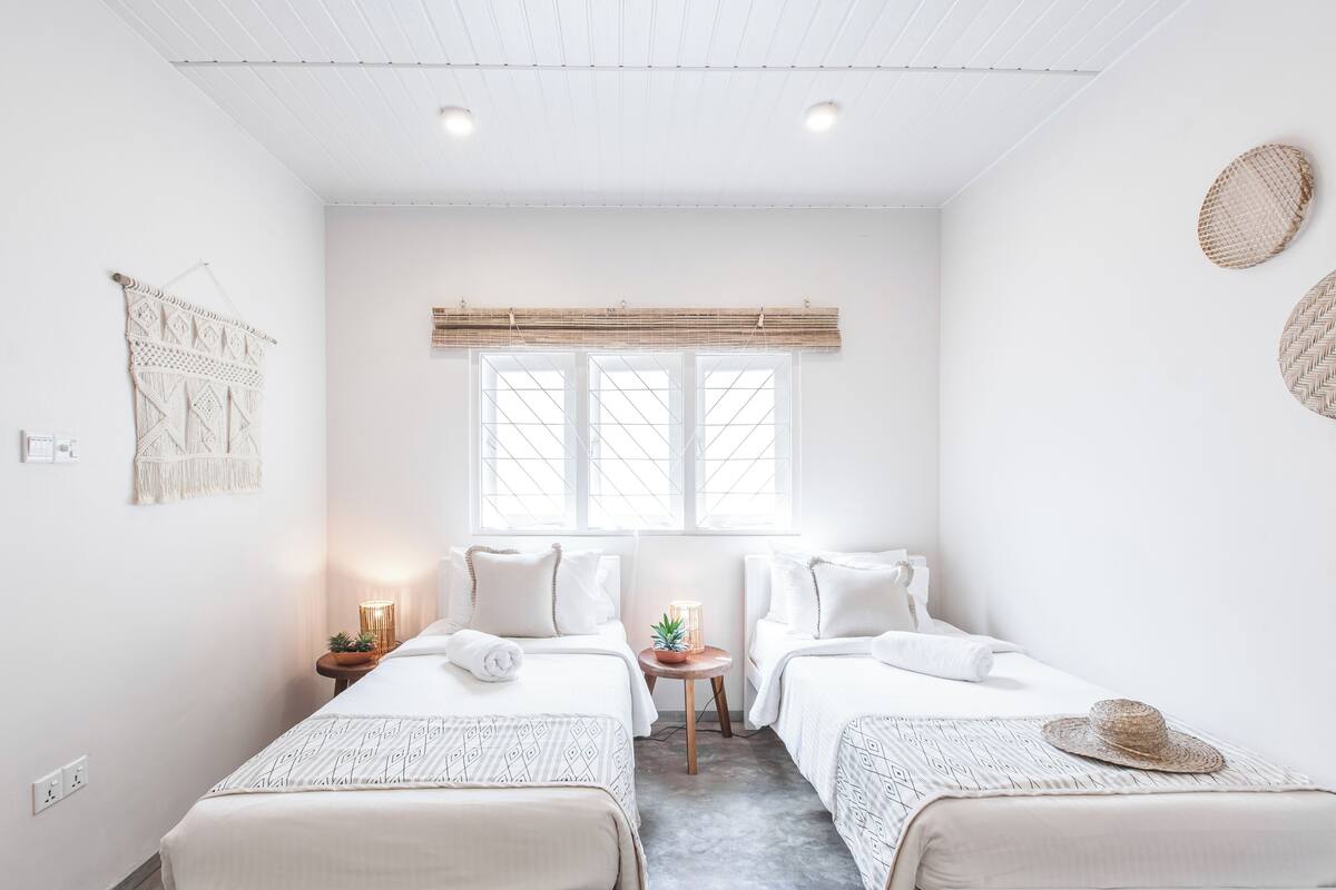 Quarto minimalista com duas camas, uma janela com persianas, uma pequena mesa com luminária e uma peça de macramé pendurada na parede.