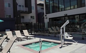 Área de piscina com espreguiçadeiras, uma banheira de hidromassagem e um edifício moderno ao fundo.