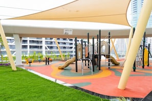 Um parquinho ao ar livre com diversas estruturas de recreação, uma área coberta e vista para um edifício moderno ao fundo.