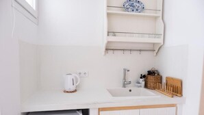 Uma pequena cozinha compacta com bancada branca, pia, chaleira e estante de madeira.