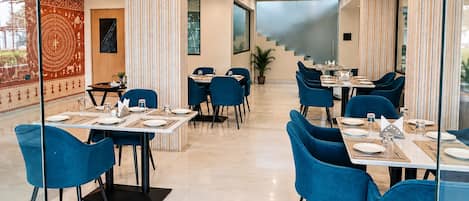 Restaurante com cadeiras azuis e mesas brancas, com uma escadaria ao fundo e uma parede com padrão.