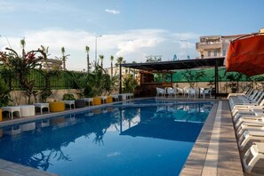 Uma piscina com espreguiçadeiras e mesas, cercada por vegetação e edifícios.