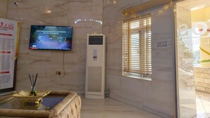 Quarto de hotel com televisão fixada na parede, janela com persianas, ar condicionado montado na parede e bandeja decorativa com castiçal.