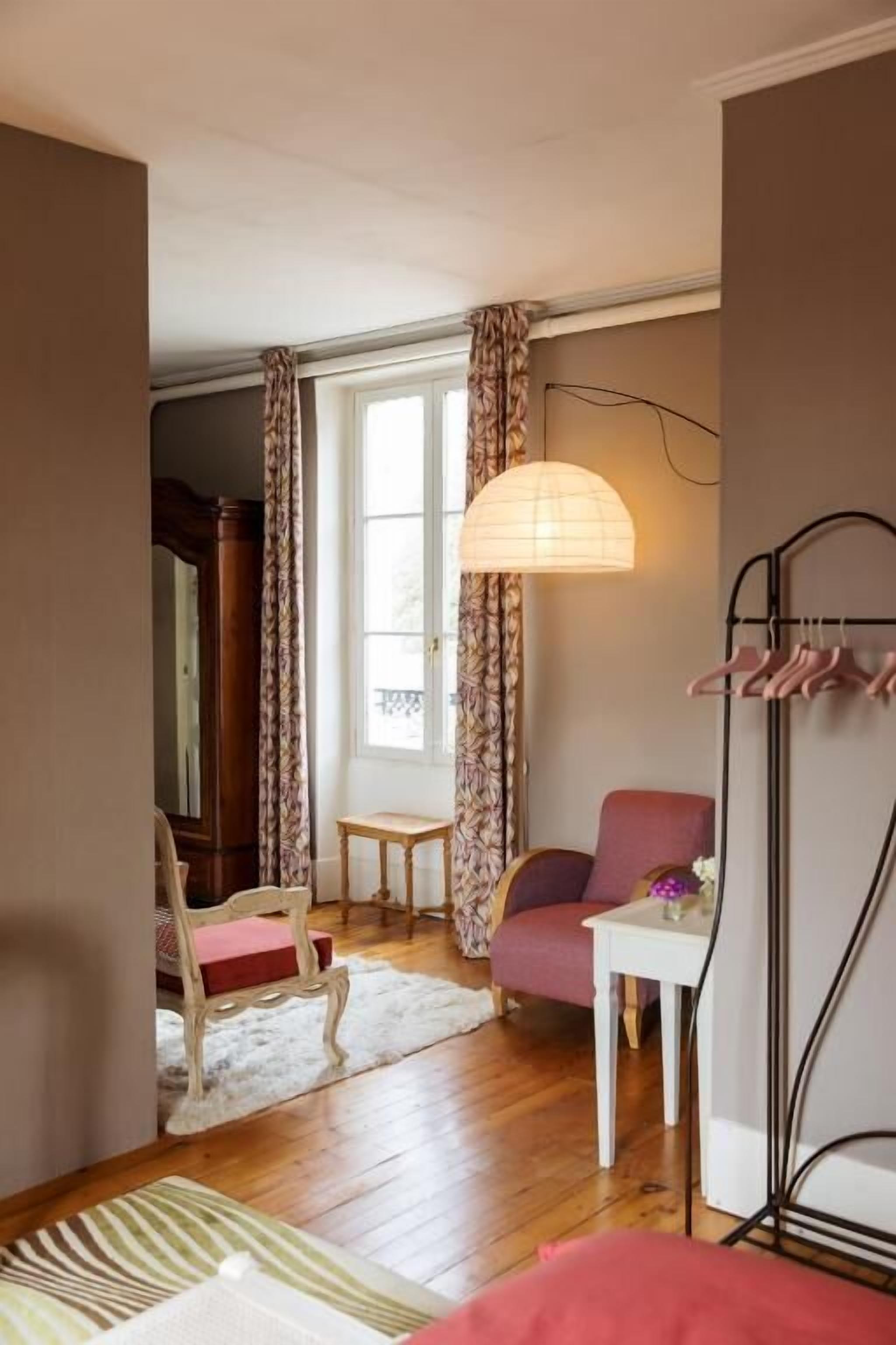 Um quarto com piso de madeira, uma cadeira rosa, uma mesa branca e um candeeiro pendente.