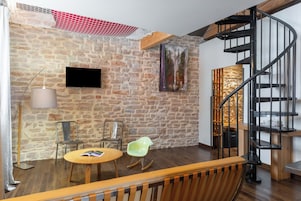 Quarto com parede de pedra, televisão de tela plana, mesa de madeira, duas cadeiras, uma cadeira verde, escada em espiral e luminária pendente.