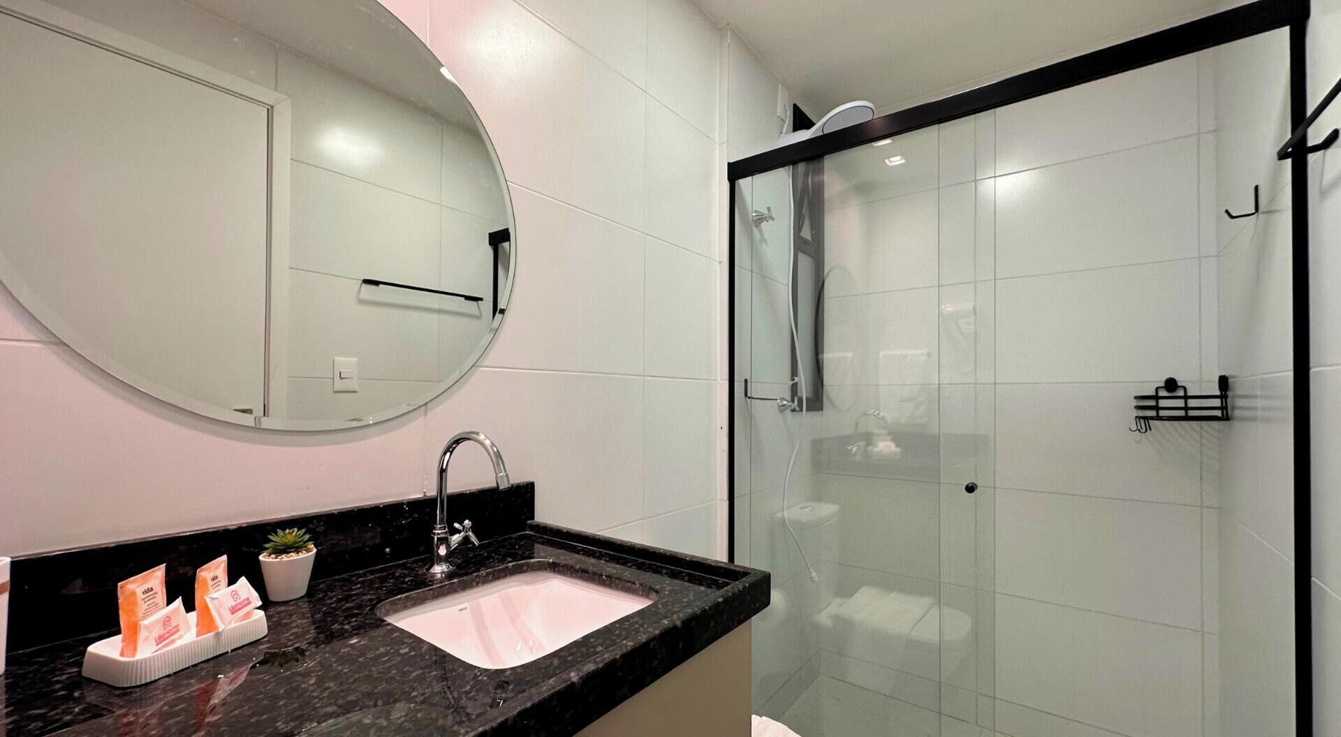 Uma casa de banho moderna com um espelho redondo, uma pia com bancada preta e cabine de duche com vidro.