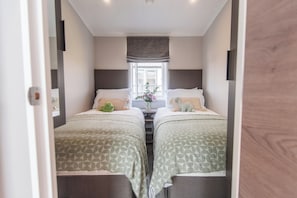 Quarto pequeno com duas camas, janela com cortinas e uma mesinha com um vaso de flores.
