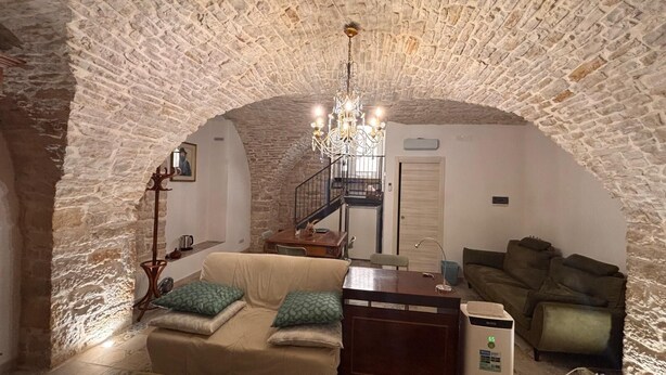 Uma sala de estar aconchegante com teto abobadado de pedra, um lustre, um sofá com almofadas, uma mesa de jantar e uma escada que leva a um andar superior.
