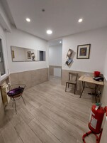 Quarto com piso de madeira, uma cadeira, uma mesinha com um objeto vermelho e um extintor de incêndio na parede, além de um espelho.