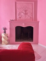 Um quarto com parede rosa, lareira com painel decorativo acima, sofá vermelho e mesa de apoio branca com abajur.