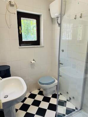 Uma casa de banho com uma pia branca, uma sanita com assento azul, uma janela com vista para vegetação e uma área de duche com porta de vidro.