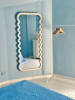 Um quarto com um espelho ondulado, um penteado branco, um tapete azul e um cabideiro com ganchos.