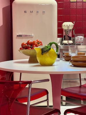 Uma cozinha retrô com uma geladeira branca, uma tigela de tomates, um jarro amarelo e cadeiras vermelhas.