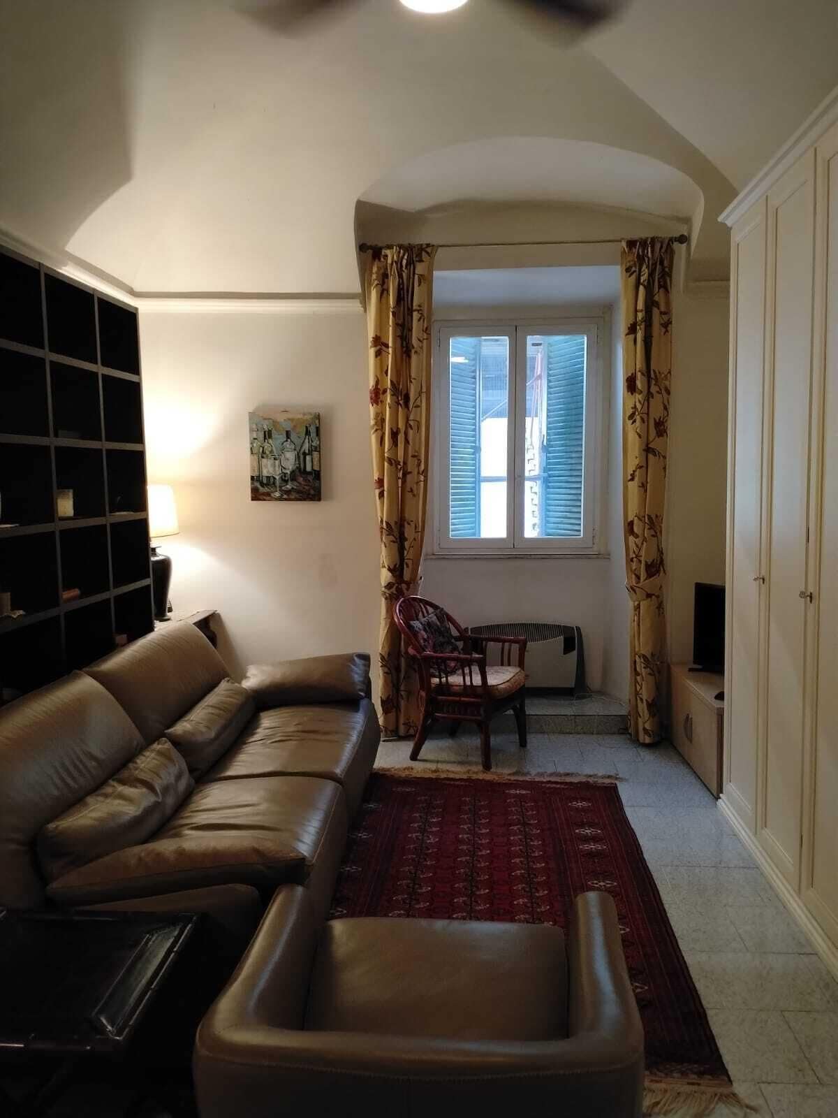Uma sala de estar com um sofá, uma poltrona, um tapete vermelho, uma estante de livros, um quadro e uma janela com cortinas.