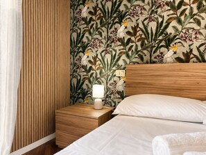 Quarto de hotel com cabeceira de madeira, mesa de cabeceira com abajur e papel de parede floral.