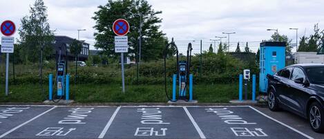 Uma área de estacionamento com estações de carregamento para veículos elétricos, vagas demarcadas e uma placa de proibido estacionar.
