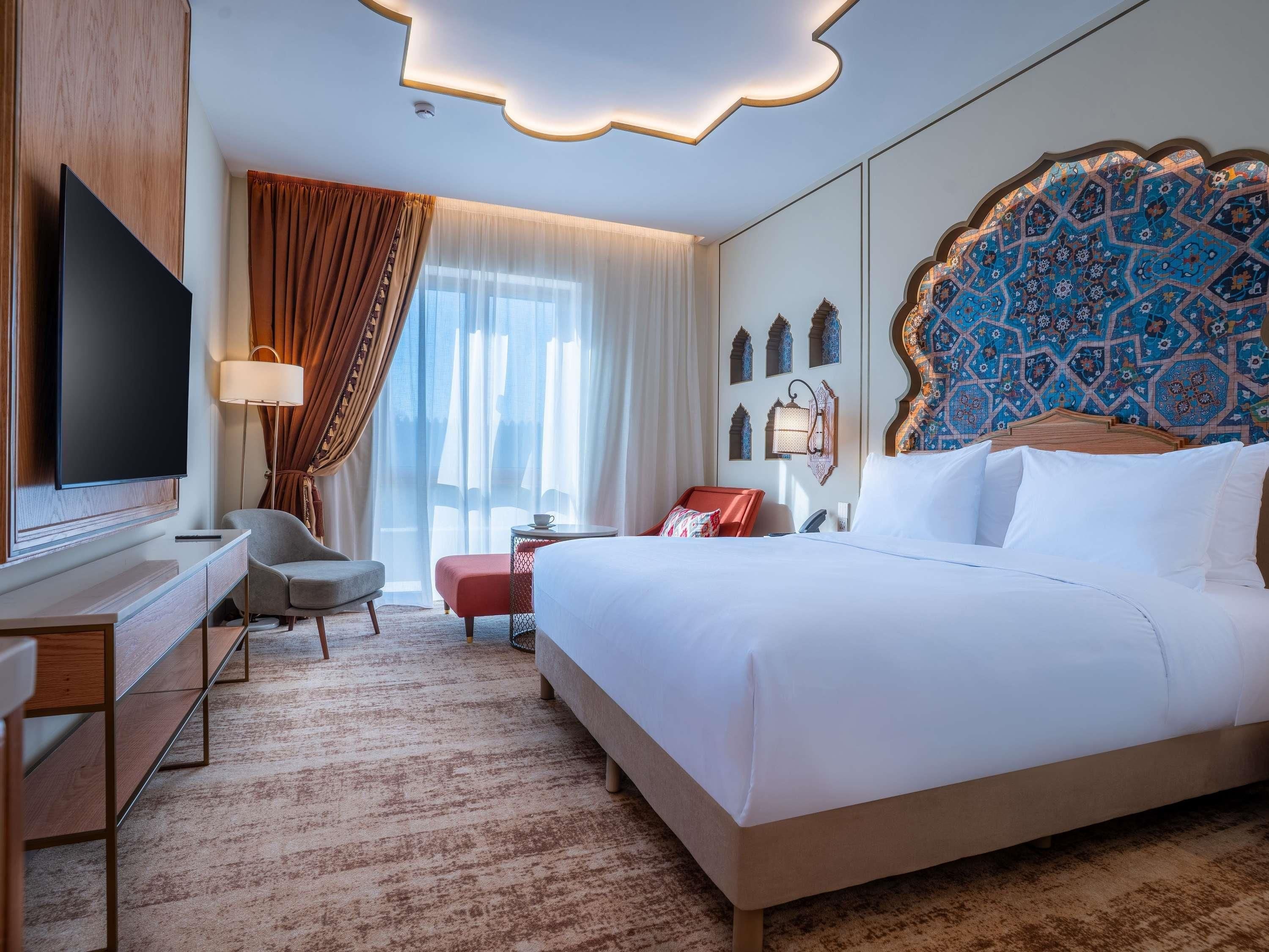 Quarto de hotel com uma cama grande, televisão de tela plana, escrivaninha com cadeira, mesa pequena e cabeceira decorativa.
