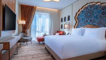 Quarto de hotel com uma cama grande, televisão de tela plana, escrivaninha com cadeira, mesa pequena e cabeceira decorativa.